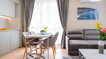 Uma sala de estar moderna com sofá, mesa de jantar e cadeiras. Há uma janela com cortinas, um quadro na parede e um vaso com flores.