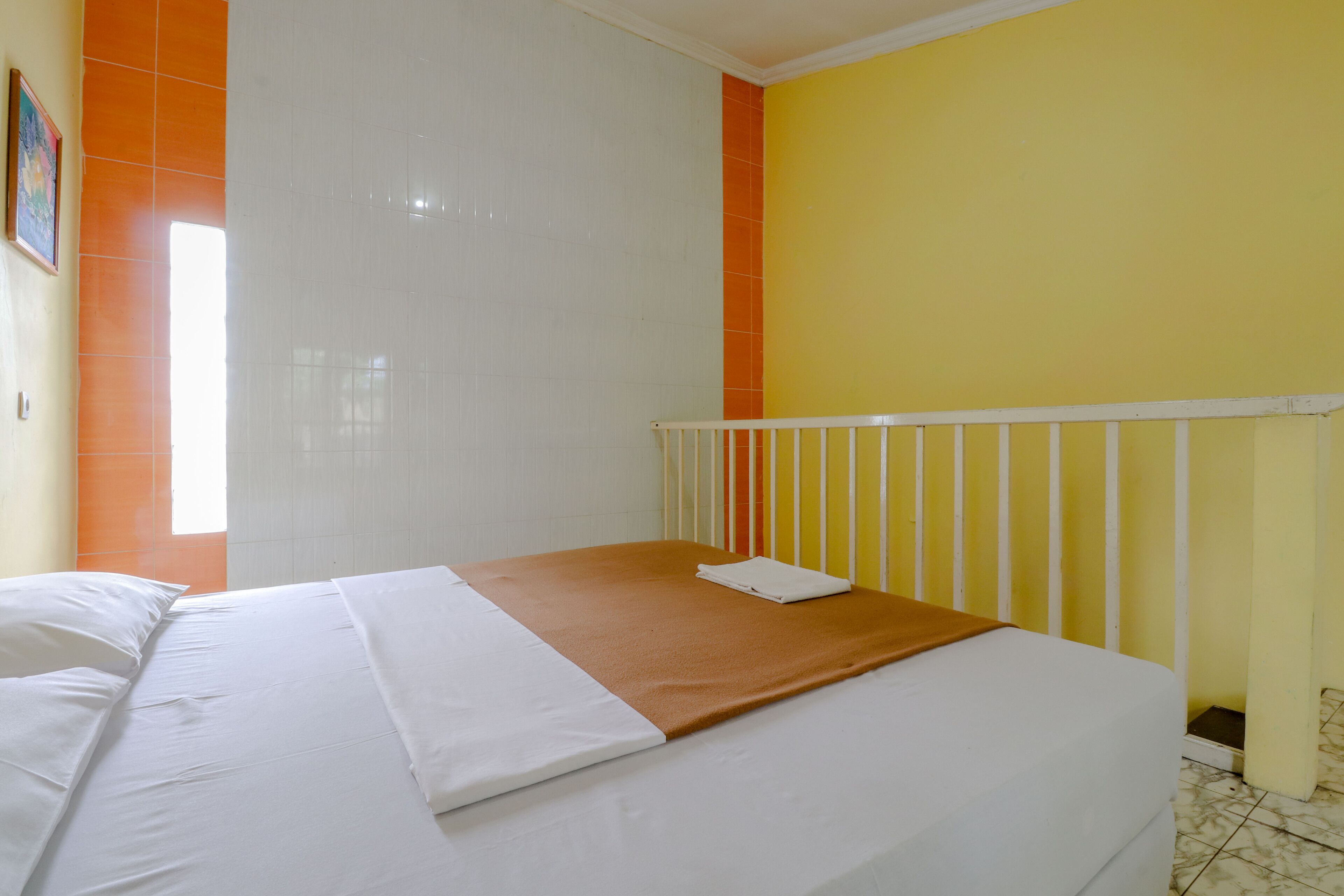 Cama bem arrumada com roupa de cama branca e castanha, cabeceira branca e parede amarela com uma janela.