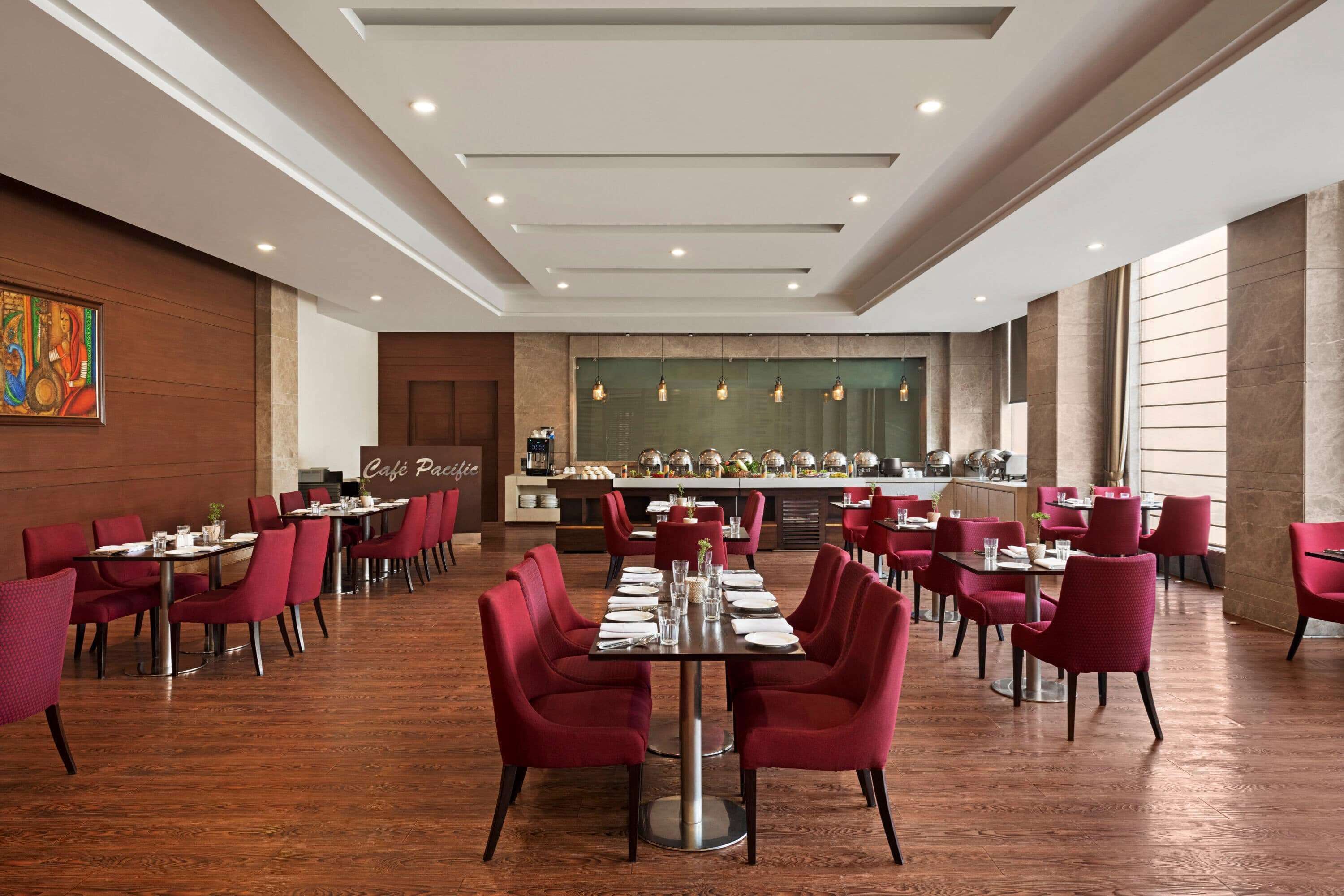 Uma ampla área de refeições com cadeiras estofadas em vermelho e mesas, um bar central e um design de teto moderno.