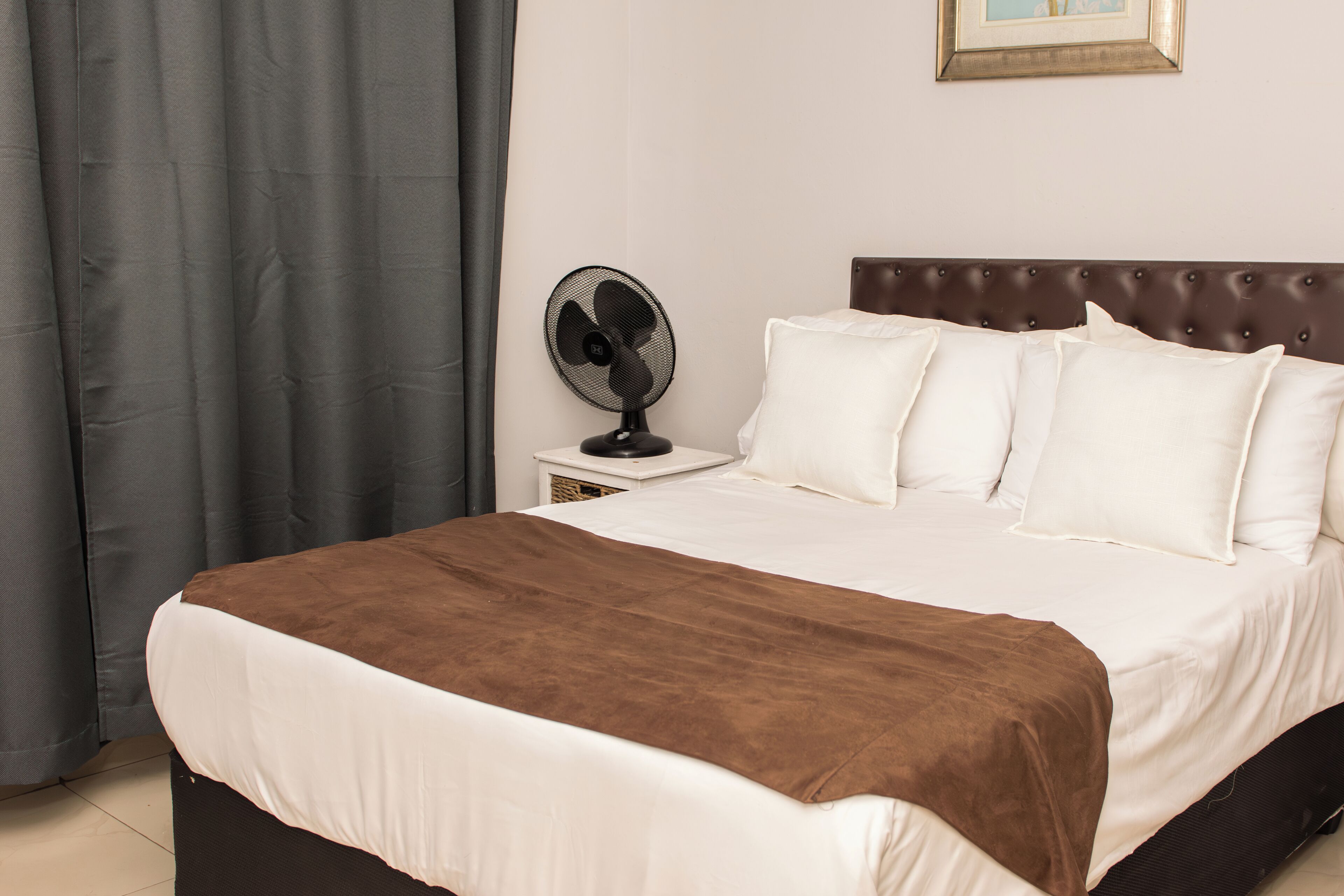 Uma cama bem arrumada com roupa de cama branca e castanha, uma mesinha de cabeceira com um ventilador e uma cortina escura à esquerda.