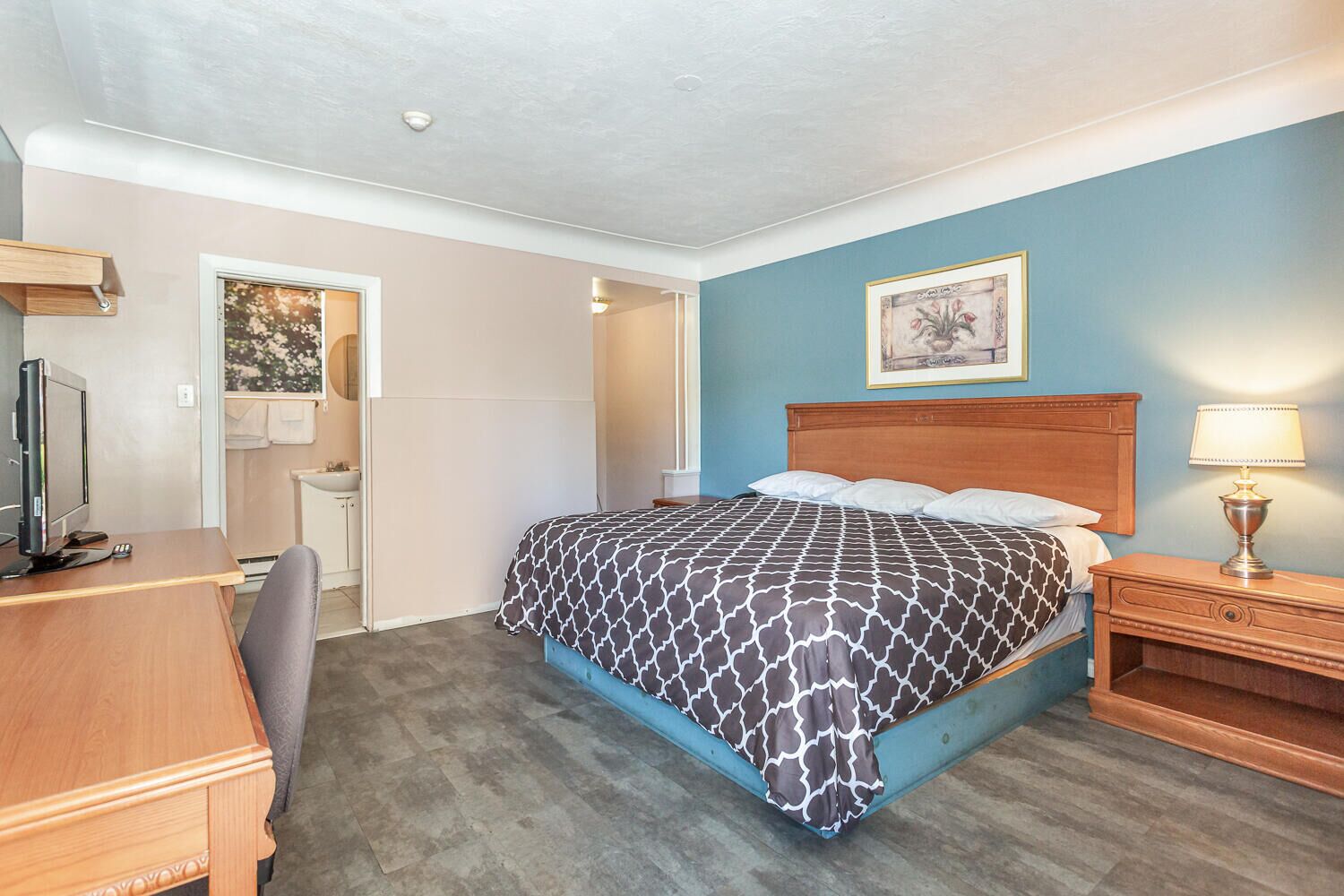 Quarto com cama, mesa de cabeceira, luminária, escrivaninha, cadeira e janela com vista para árvores.