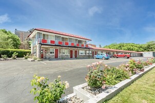 Um motel com telhado vermelho e vários quartos, estacionamento e um jardim com flores.