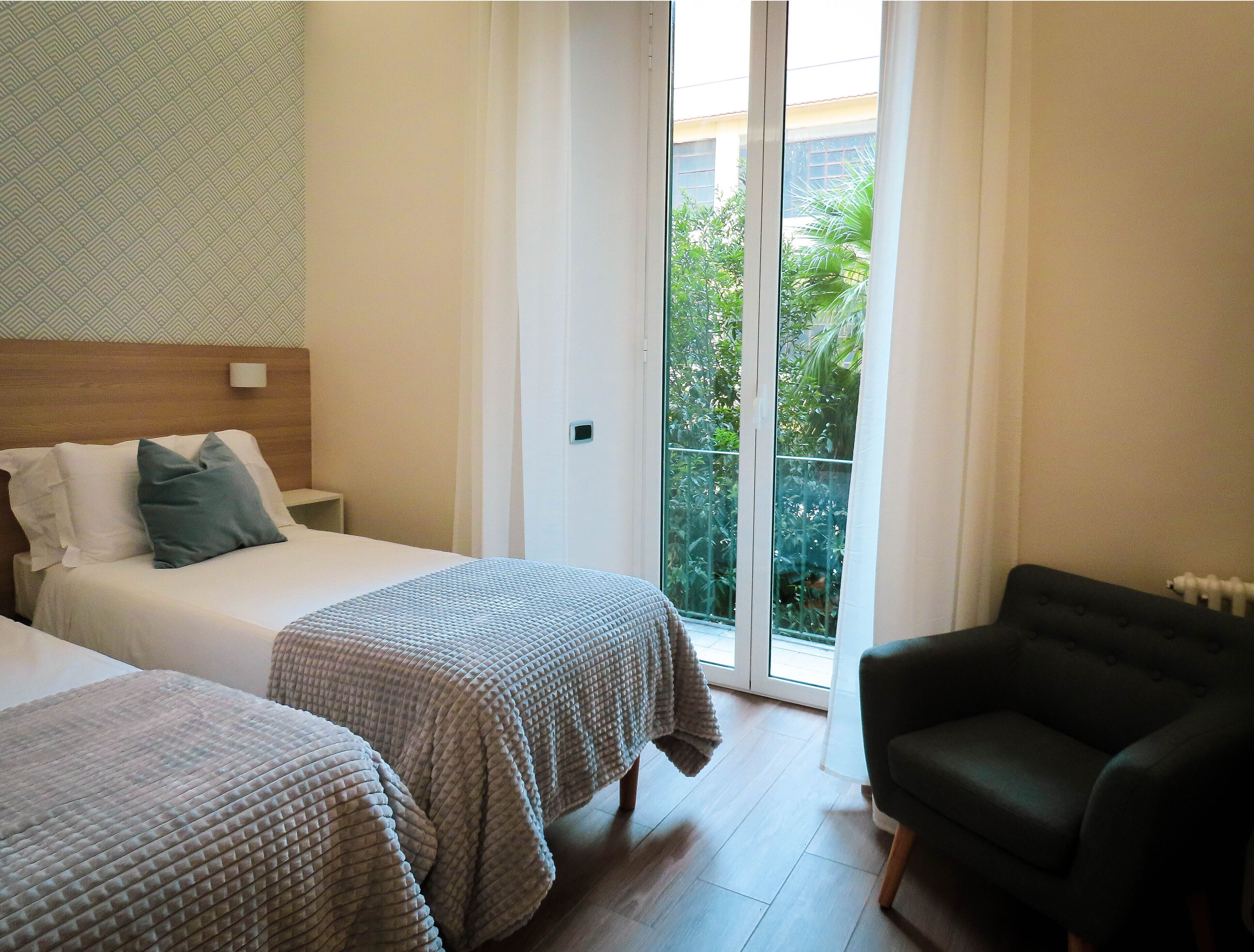 Quarto de hotel com duas camas, uma poltrona escura e vista para o verde pela janela.