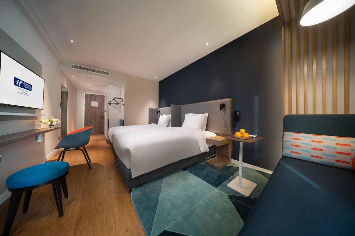 Quarto de hotel moderno com uma cama grande, uma escrivaninha, uma cadeira e uma pequena mesa com frutas.