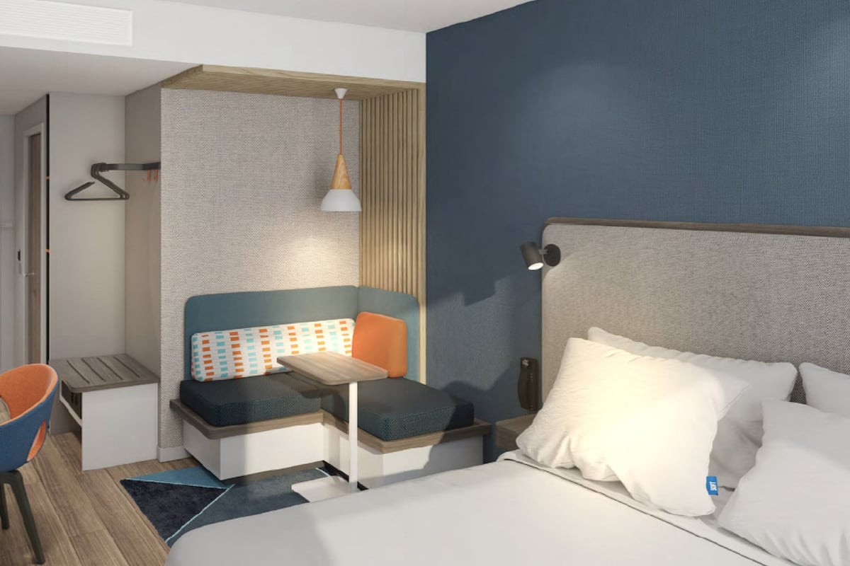 Quarto de hotel compacto com cama, pequena mesa de trabalho, cadeira e área de estar com almofadas.