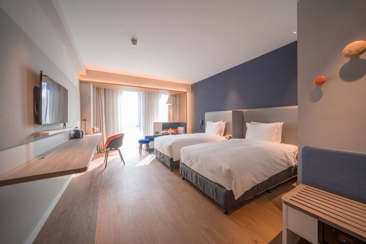 Quarto de hotel moderno com duas camas, uma escrivaninha, uma cadeira e uma televisão.