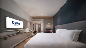 Quarto de hotel moderno com uma cama grande, televisão de tela plana na parede, escrivaninha com cadeira e uma mesinha com um vaso de flores.