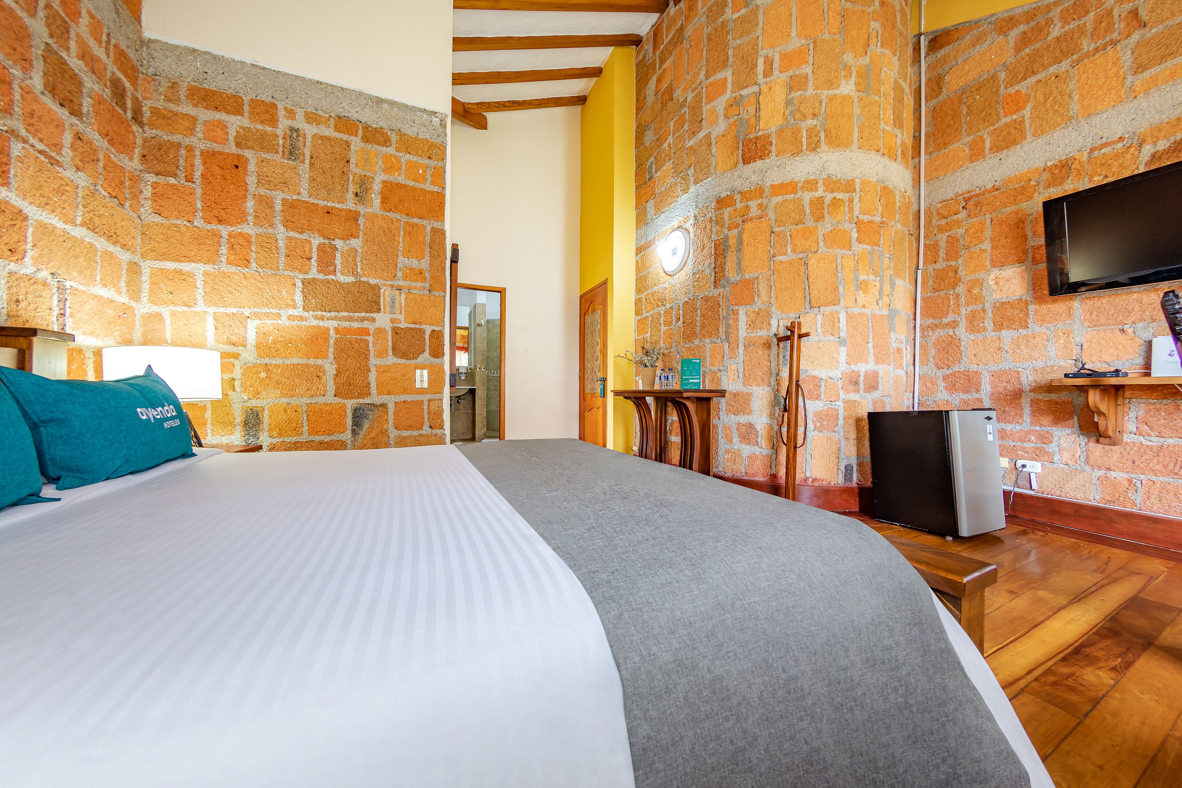 Quarto de hotel com parede de tijolos, uma cama com cobertor cinza, TV na parede e uma pequena geladeira.
