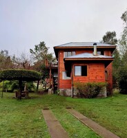 Uma cabana de madeira de dois andares com varanda, cercada por um gramado e árvores.