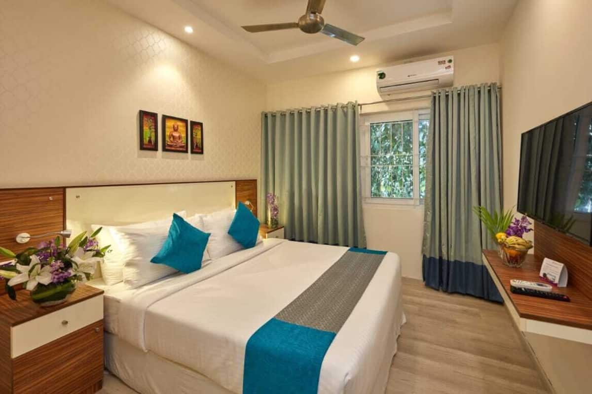 Quarto de hotel com cama, escrivaninha, espelho e janela com cortinas.