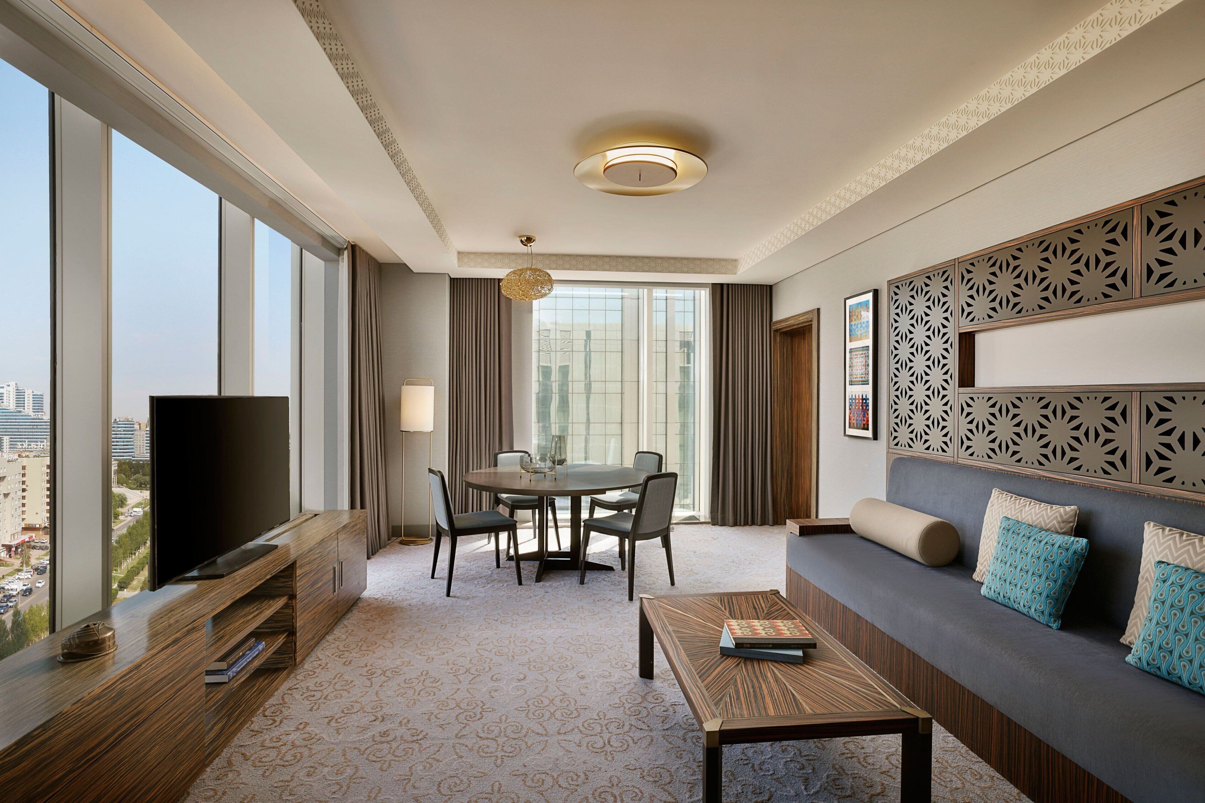 Quarto de hotel moderno com uma janela ampla, televisão de tela plana, mesa de jantar com cadeiras, sofá com almofadas estampadas e um painel decorativo na parede.
