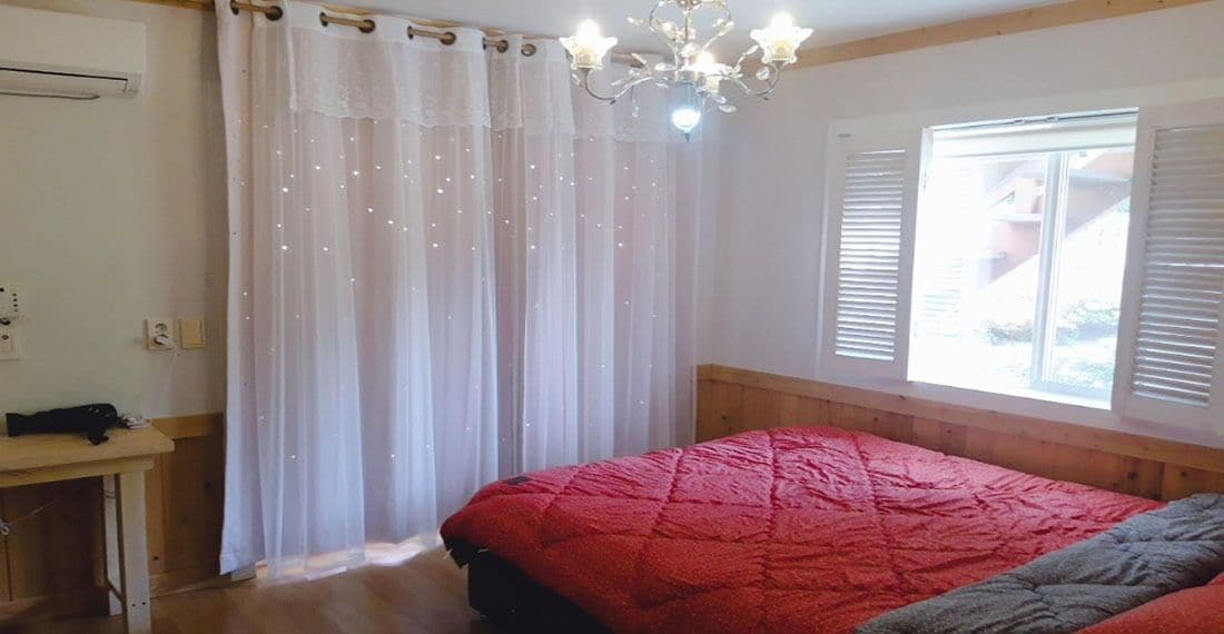 Quarto com colcha vermelha, cabeceira de madeira, cortina branca e lustre.