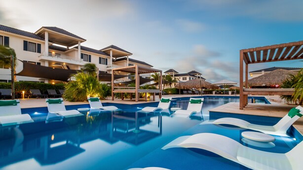 Piscina com espreguiçadeiras, área de estar coberta e prédio de resort com várias varandas.