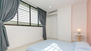 Quarto com cama, cortinas, janela com persianas, ar condicionado e uma mesinha de cabeceira com abajur.