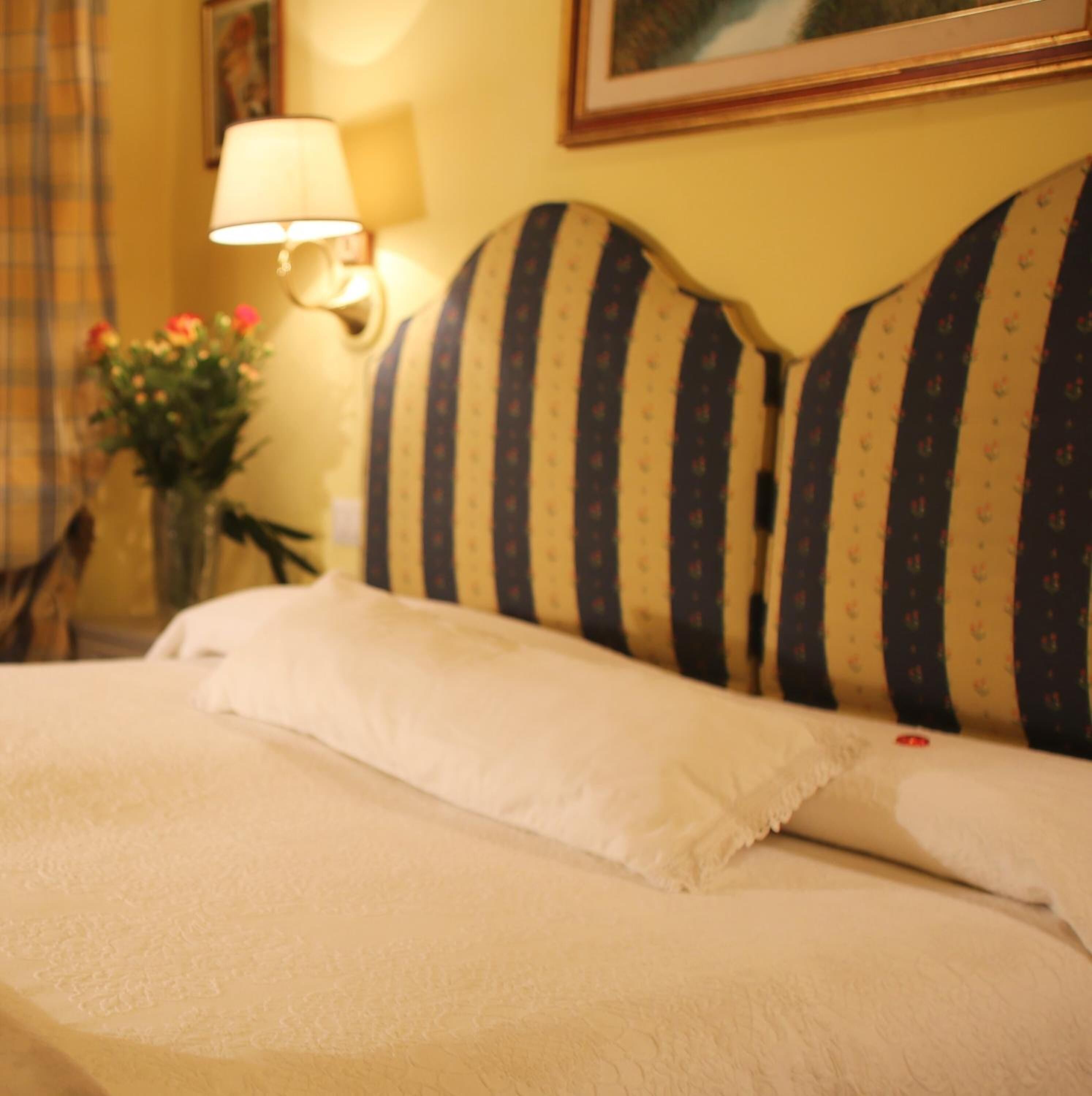 Quarto de hotel com cama, almofadas, abajur e um vaso com flores.