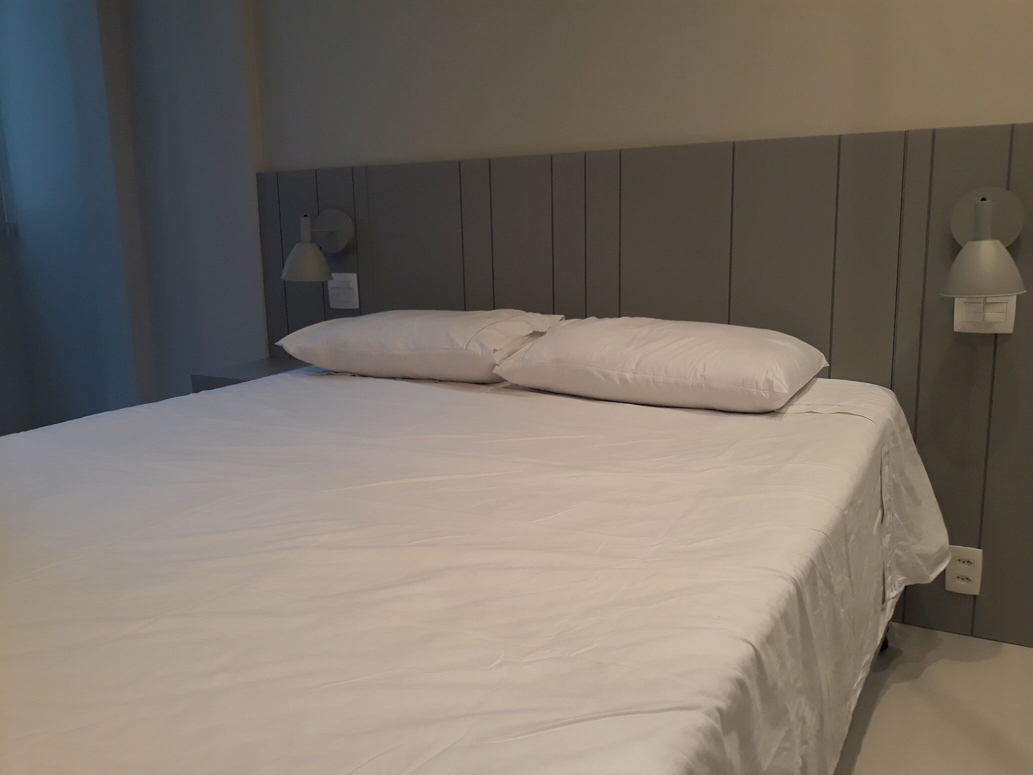 Cama bem arrumada com roupa de cama e almofadas brancas, uma luminária na parede e uma tomada.