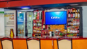 Uma área de bar com uma tela de televisão exibindo 'DSTV', prateleiras repletas de garrafas de bebidas alcoólicas e um anúncio de 'New Tusker Premium Ale'.