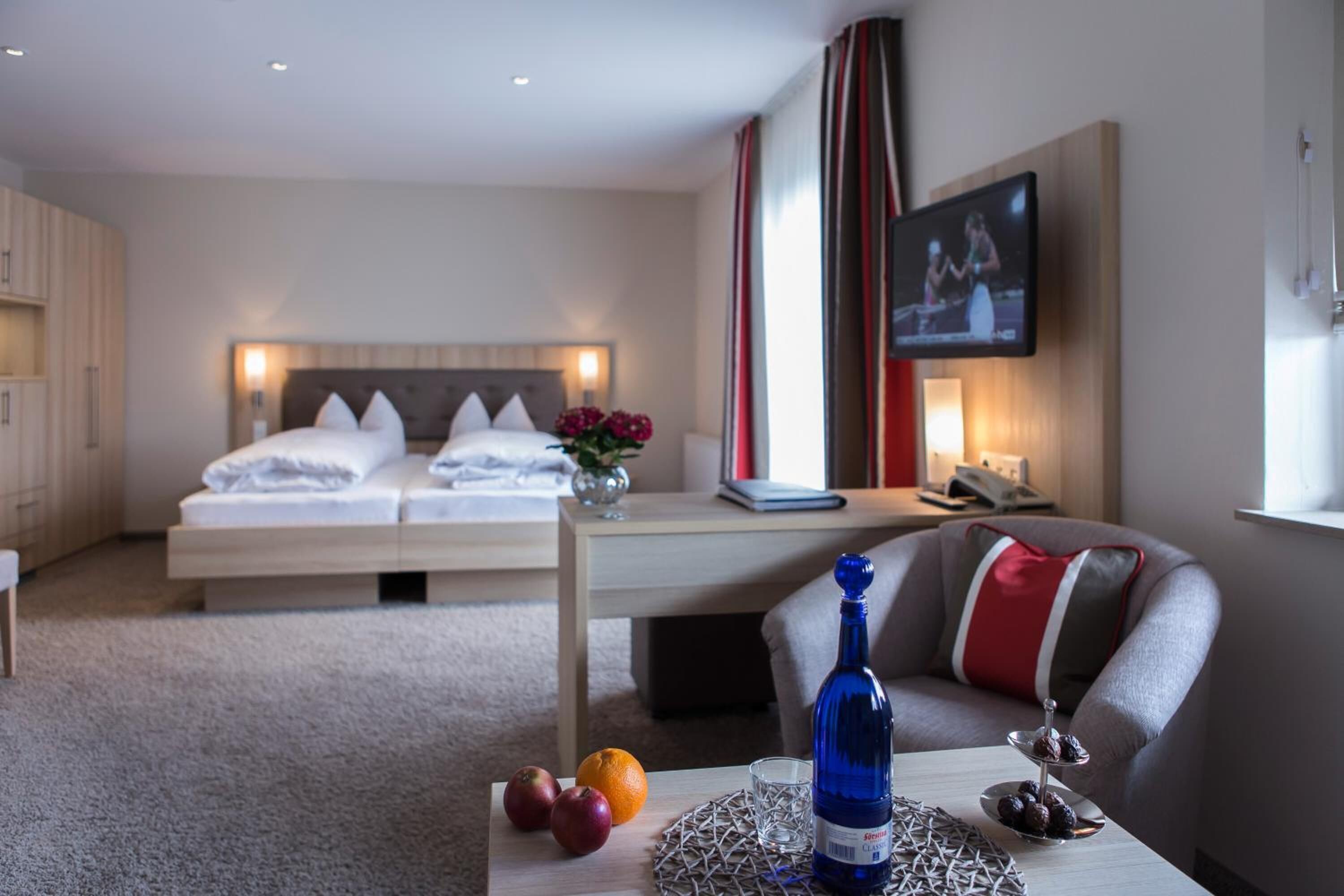 Quarto de hotel com cama, televisão, sofá, mesa com frutas e garrafa, e janela com cortinas.