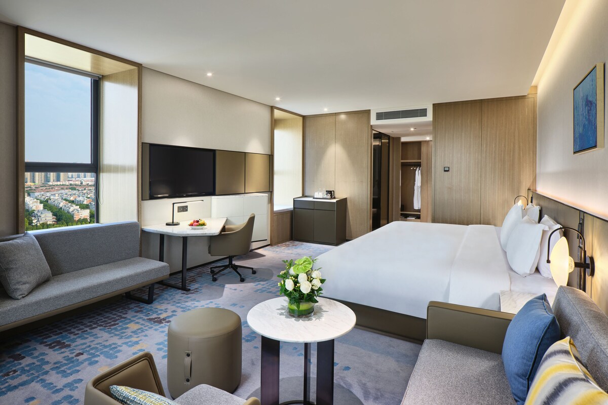 Quarto de hotel moderno com uma cama grande, um sofá, uma escrivaninha e vista para a cidade.