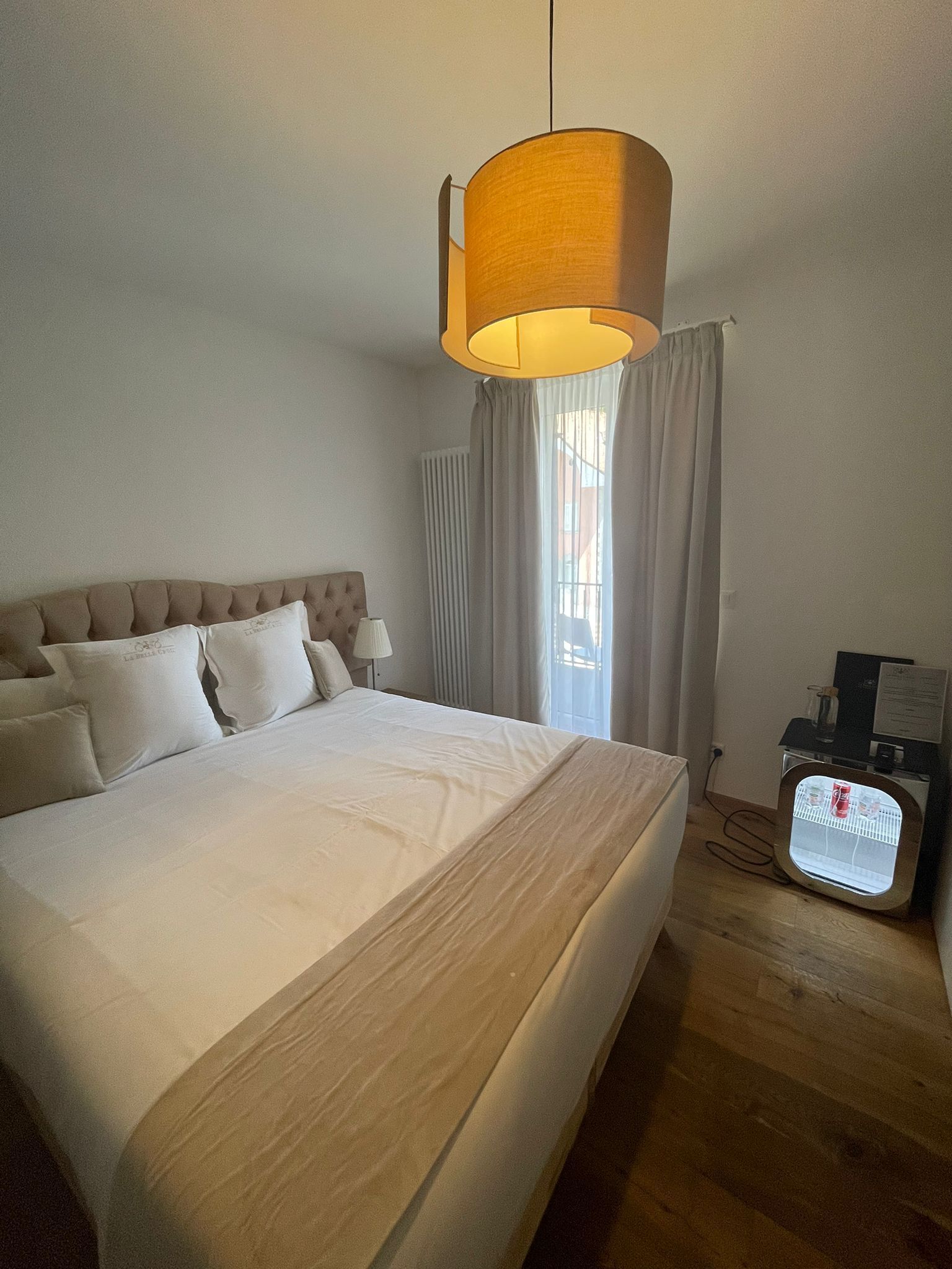 Cama bem arrumada com cabeceira, luminária de cabeceira e janela com cortinas.