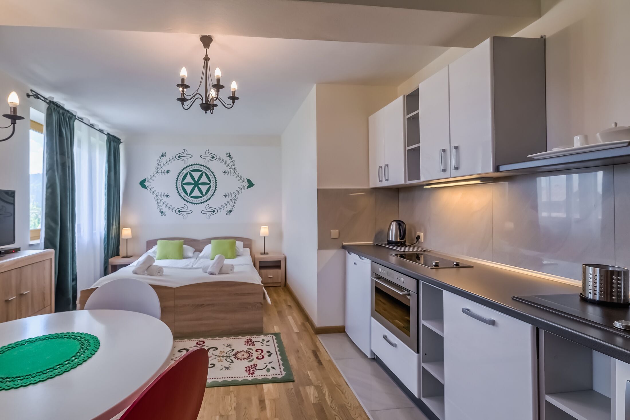 Quarto de hotel moderno com cama, mesas de cabeceira, área de refeições e cozinha compacta.