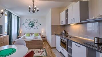 Quarto de hotel moderno com cama, mesas de cabeceira, área de refeições e cozinha compacta.