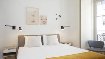 Quarto de hotel com uma cama grande, mesas de cabeceira, uma cadeira, uma obra de arte na parede e uma janela.