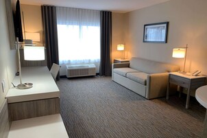 Quarto de hotel com cama, escrivaninha com luminária, sofá, mesa de cabeceira e janela com cortinas.