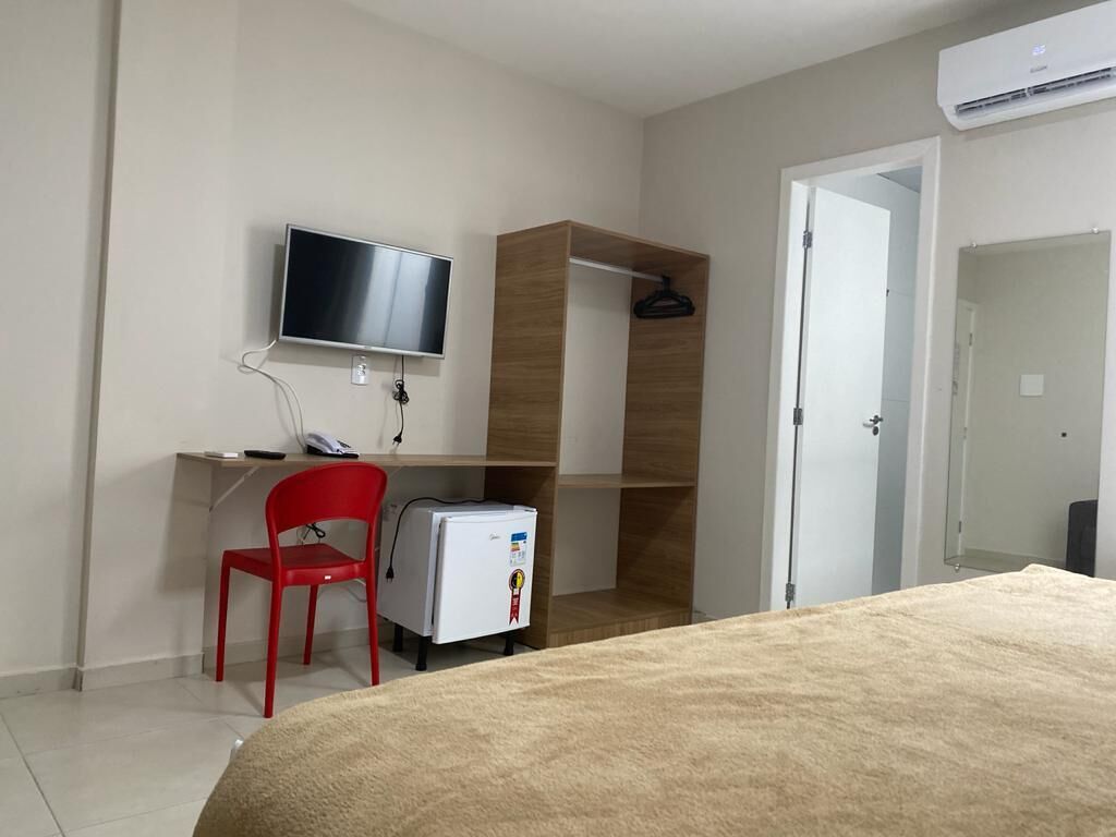 Quarto com cama, escrivaninha com televisão, cadeira vermelha, guarda-roupa e um pequeno frigorífico.