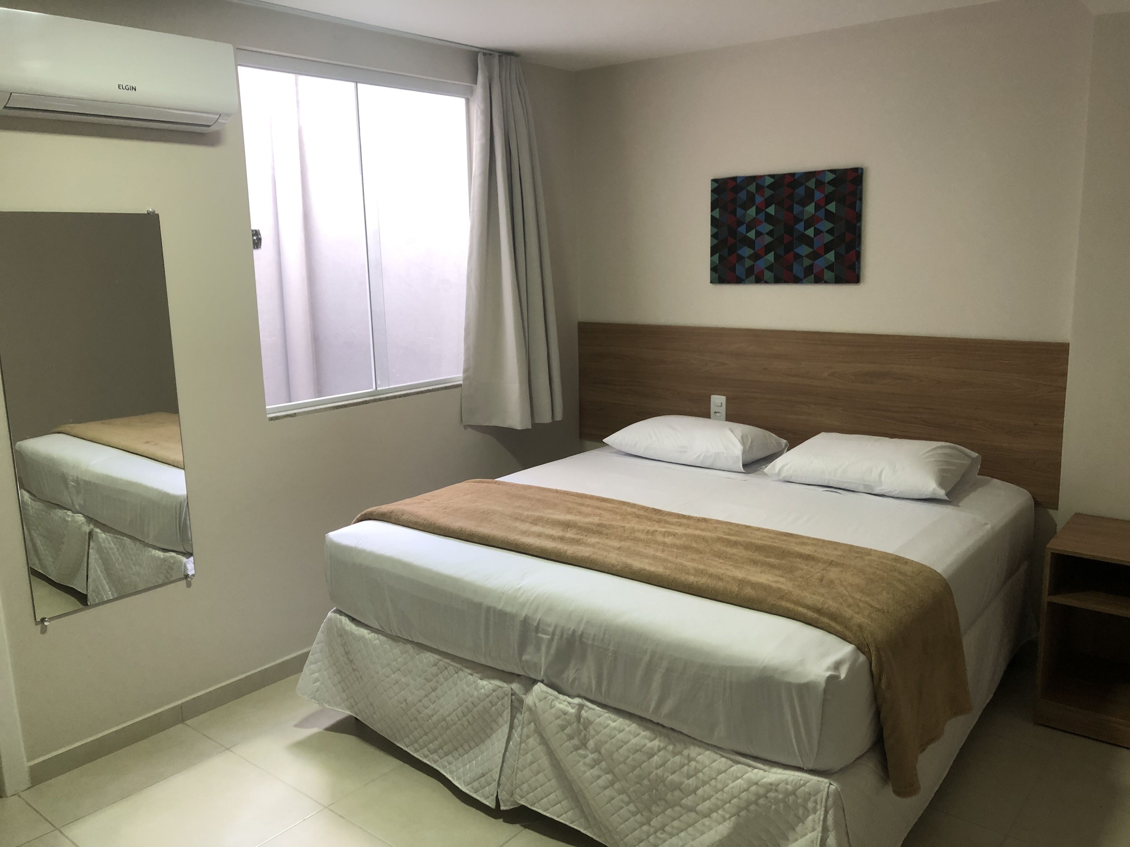 Quarto de hotel com cama, janela com cortinas, ar-condicionado fixado na parede, espelho e uma pequena mesa de cabeceira de madeira.