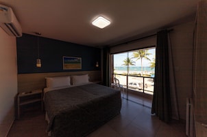 Quarto de hotel com uma cama grande, uma mesa de cabeceira, uma janela vista para a praia e uma luminária no teto.