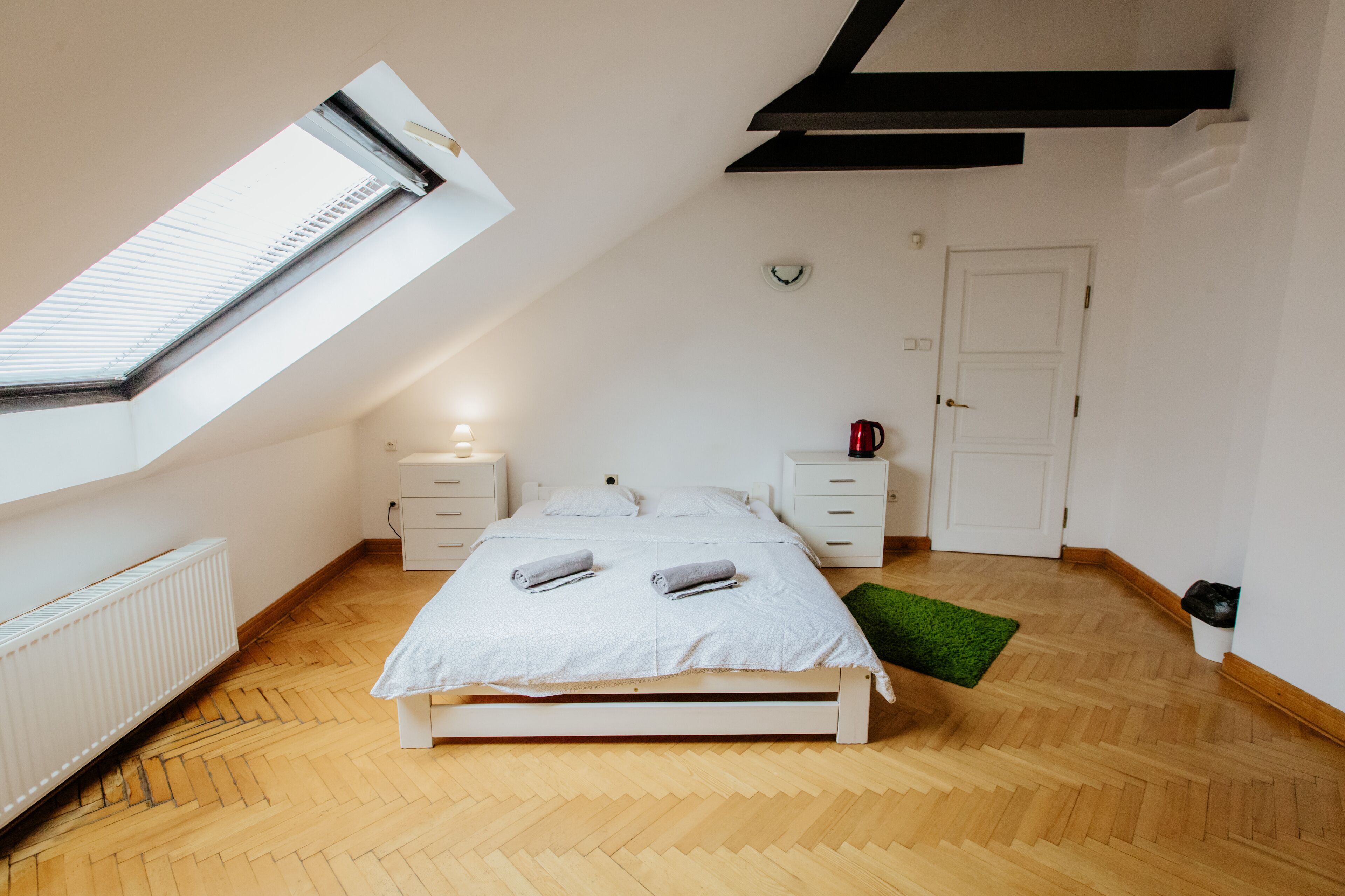 Quarto com clarabóia, piso de madeira, cama branca, mesas de cabeceira e uma porta.