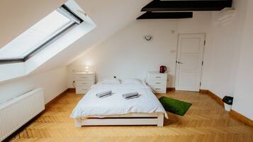 Quarto com clarabóia, piso de madeira, cama branca, mesas de cabeceira e uma porta.