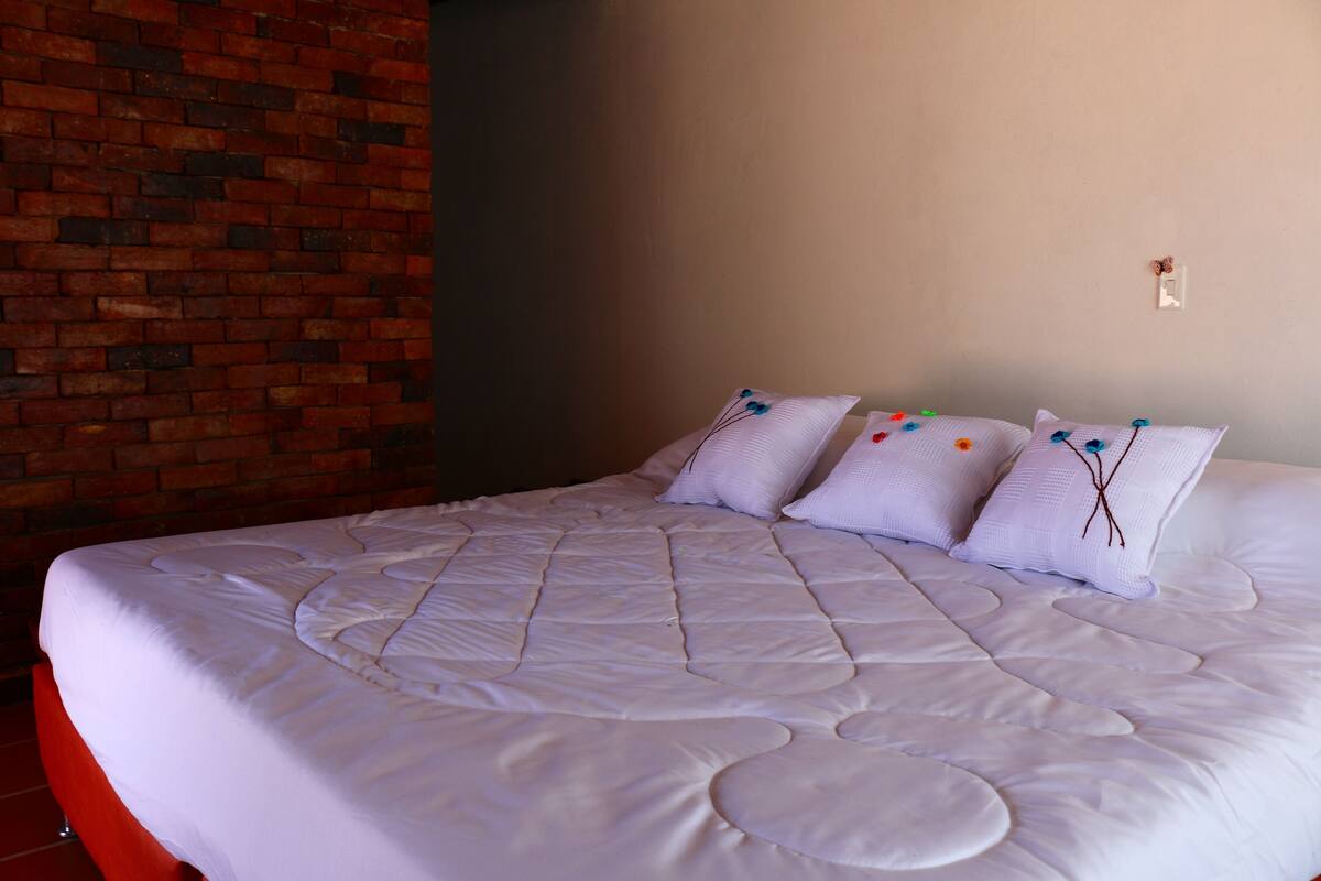 Uma cama bem arrumada com colcha e travesseiros brancos, uma parede de tijolos e uma parede lisa com um interruptor de luz.