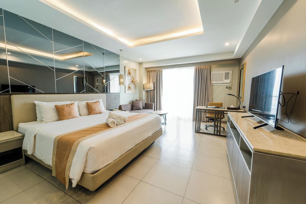 Quarto de hotel moderno com uma cama grande, uma escrivaninha, televisão e uma janela ampla com cortinas.