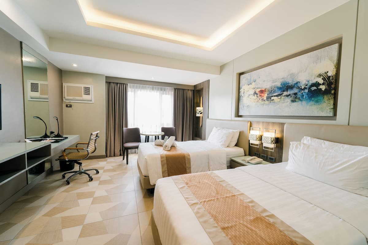 Quarto de hotel com duas camas, uma escrivaninha com computador, uma cadeira, uma luminária de mesa e um quadro na parede.