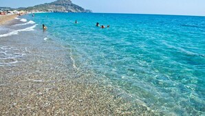 Uma praia com águas turquesa cristalinas, areal pedregoso e uma montanha ao longe.