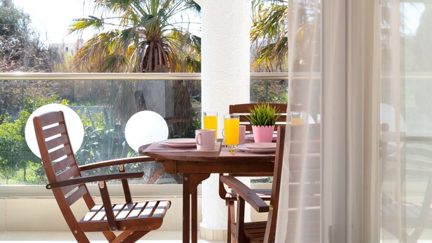 Uma mesa de madeira com cadeiras, um vaso de planta e dois copos de suco de laranja numa varanda com vista para palmeiras.