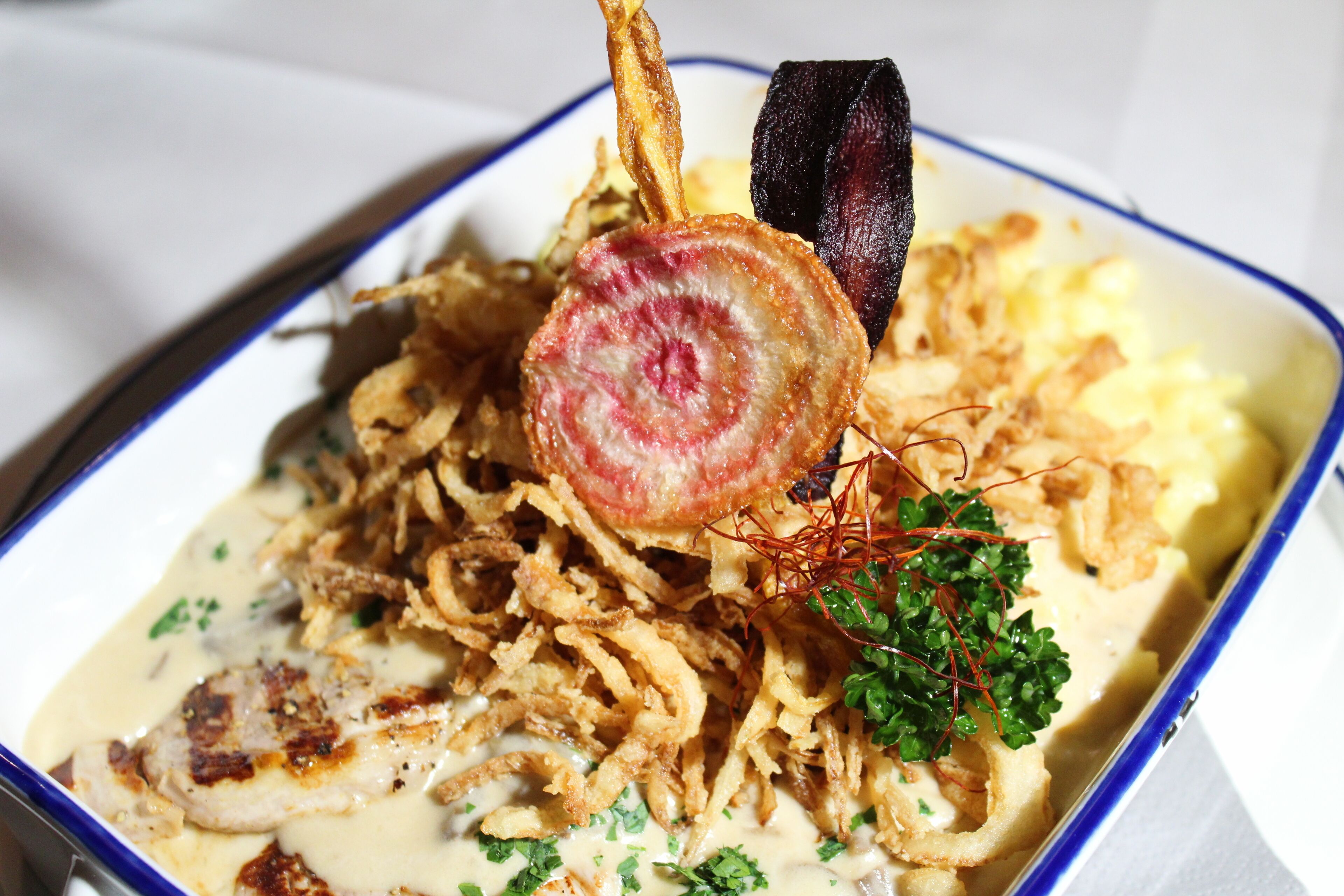 Prato com diversos ingredientes, incluindo uma fatia de carne, cebolas fritas e um enfeite.