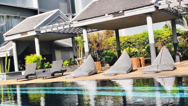Uma área à beira da piscina com espreguiçadeiras e almofadas listradas, um espaço de estar coberto e um reflexo na água.