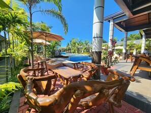 Uma área ao lado da piscina com cadeiras e mesas de madeira, cercada por palmeiras e um céu azul límpido.