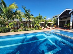 Piscina com espreguiçadeiras e guarda-sóis, cercada por palmeiras e um edifício com telhado inclinado.