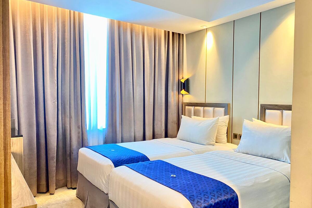 Quarto de hotel com duas camas, uma janela grande com cortinas, uma luminária fixada na parede e uma mesinha de cabeceira de madeira.