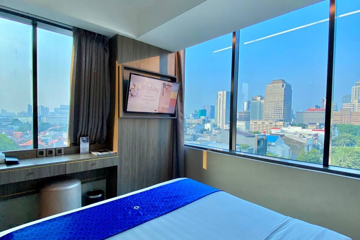 Quarto de hotel com uma cama grande, uma escrivaninha com computador, uma televisão na parede e vista para a cidade através de janelas amplas.