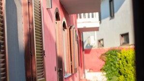 Um corredor exterior estreito, com um edifício vermelho, uma varanda com balaústres brancos e uma mesa com cadeiras.