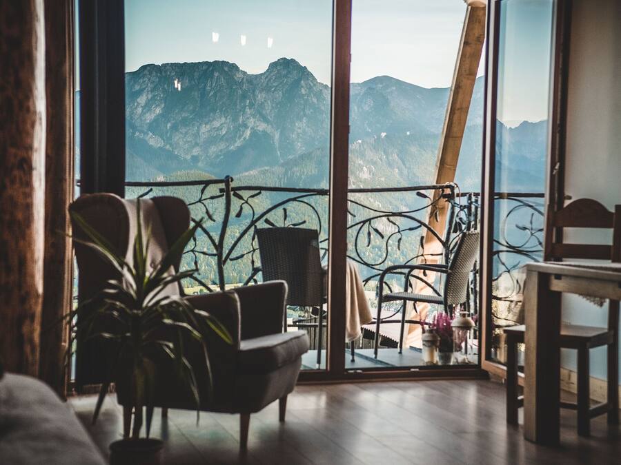 Quarto com uma grande janela que oferece vista para as montanhas, uma cadeira, uma mesa e um vaso com planta.