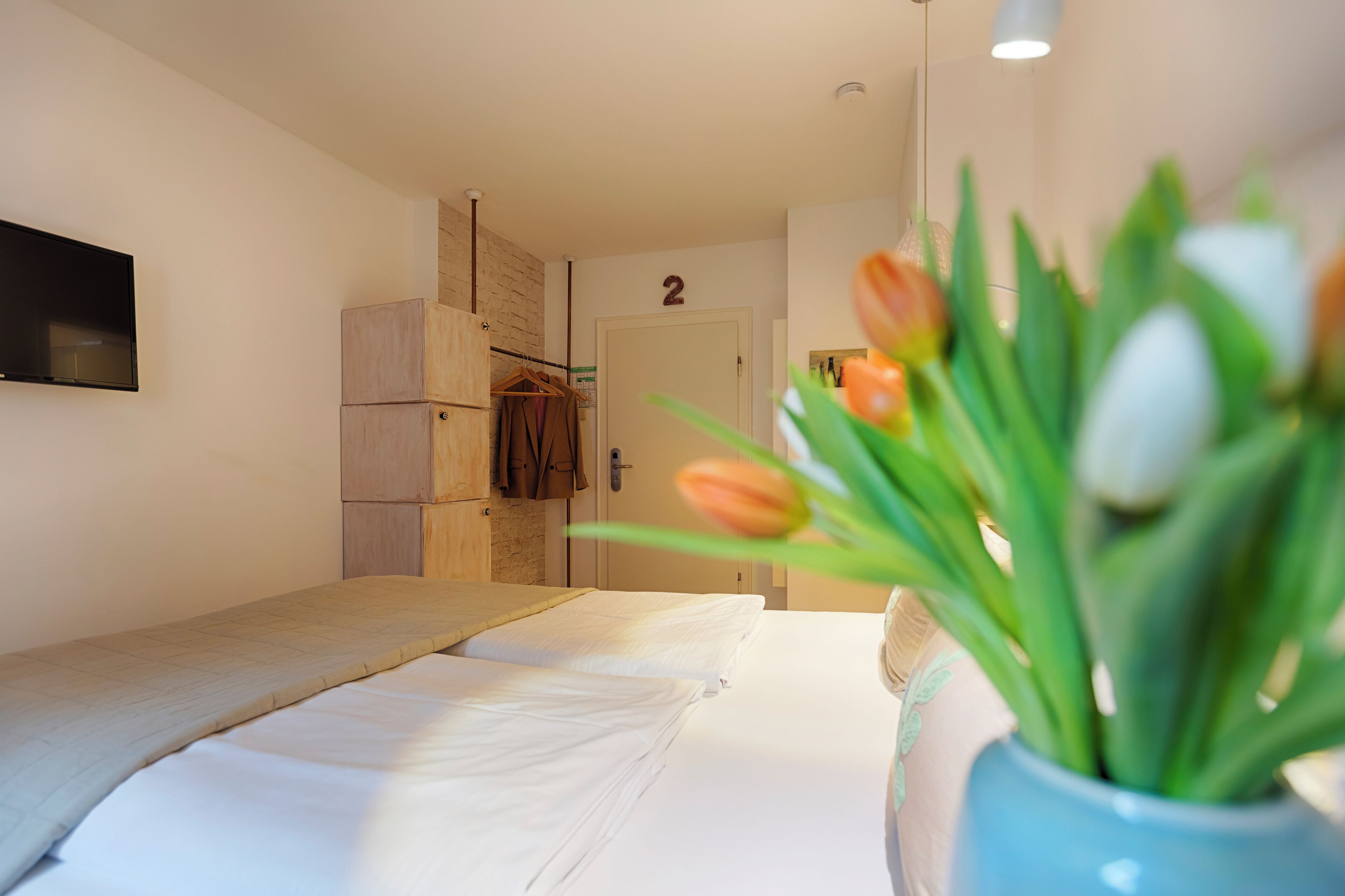 Quarto de hotel com uma cama, televisão fixada na parede, porta com o número 2 e um vaso de tulipas.