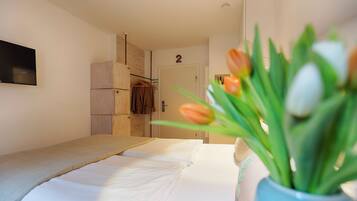 Quarto de hotel com uma cama, televisão fixada na parede, porta com o número 2 e um vaso de tulipas.