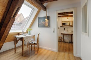 Uma sala de jantar com mesa e cadeiras, uma janela com vista, e uma cozinha visível através de uma porta aberta.