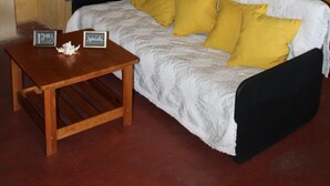 Um quarto acolhedor com teto de madeira, um sofá branco com almofadas amarelas, uma pequena mesa de madeira e uma luminária fixada na parede.